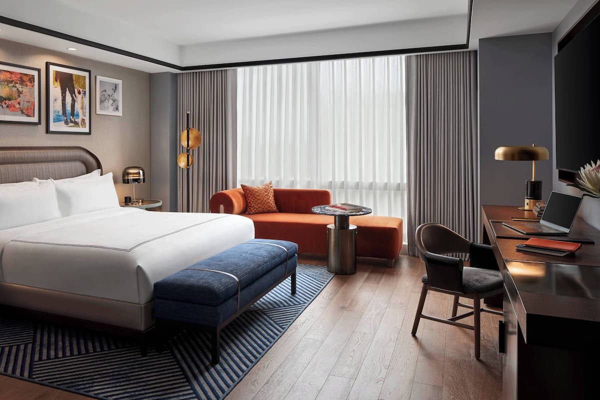 Quarto de hotel moderno com uma cama grande, uma escrivaninha com um laptop, uma cadeira, um sofá laranja e uma mesa redonda pequena.