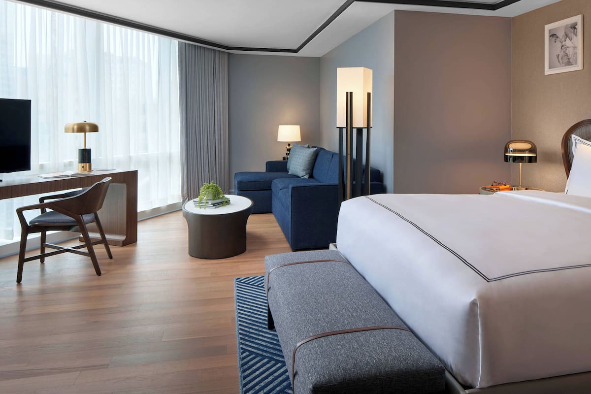 Quarto de hotel com uma cama grande, uma escrivaninha com cadeira, um sofá, uma mesinha redonda com abajur e uma televisão.