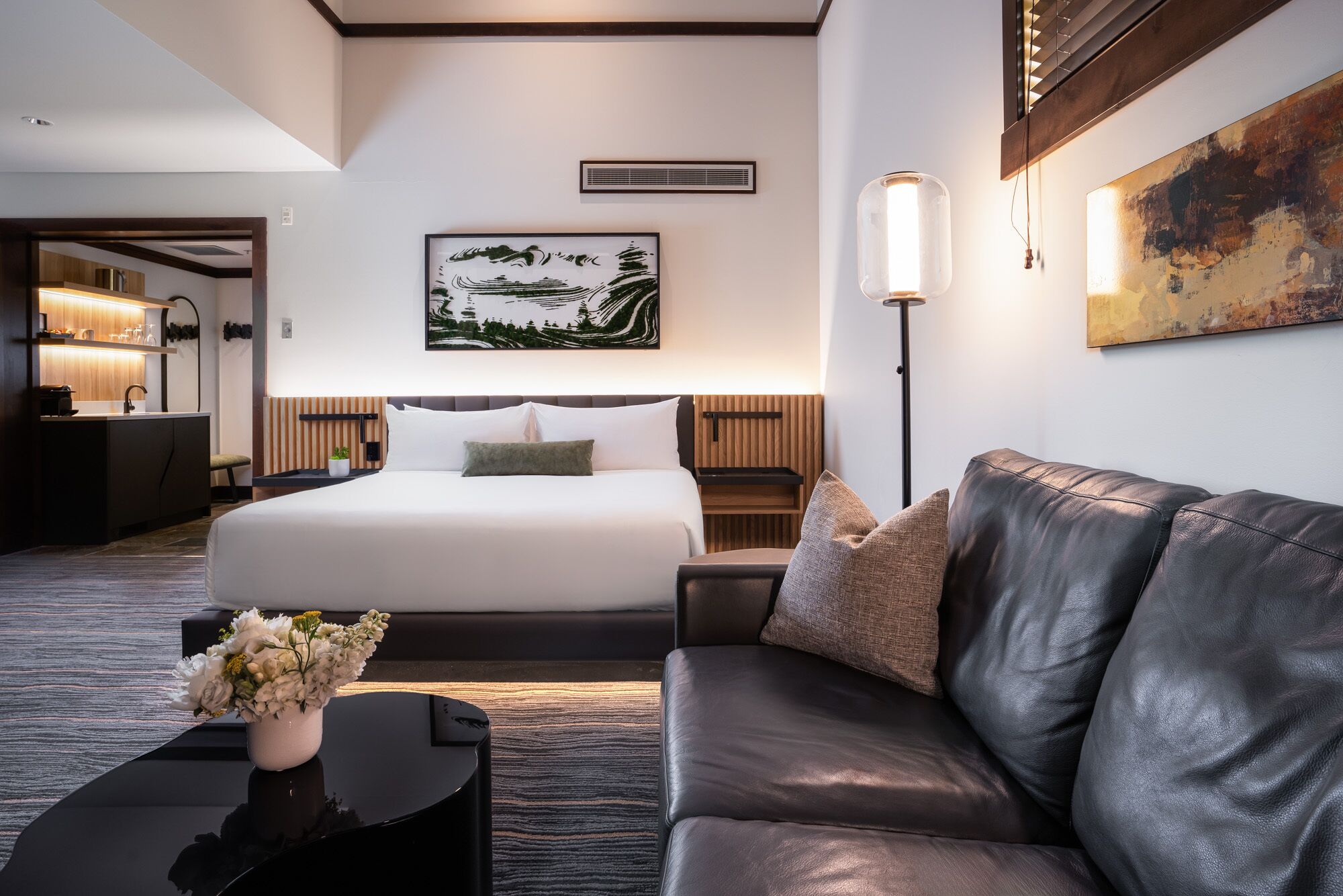 Quarto de hotel moderno com uma cama grande, um sofá de couro, uma pequena mesa com um vaso de flores e obras de arte nas paredes.