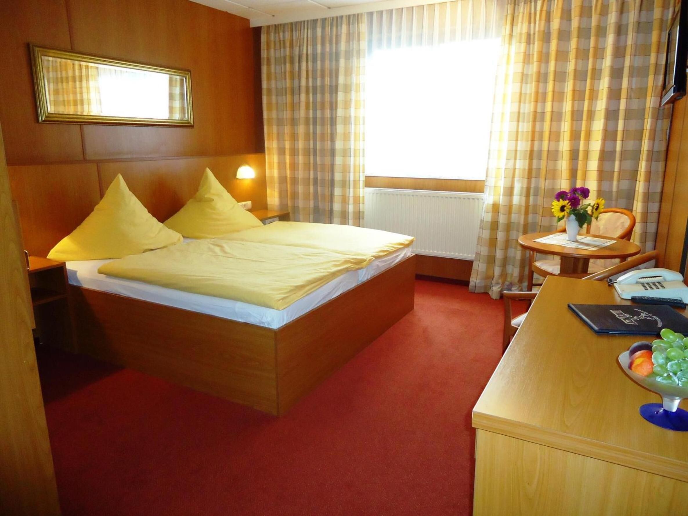 Quarto de hotel com cama, escrivaninha, cadeira, luminária, janela com cortinas e uma mesinha com um vaso de flores.