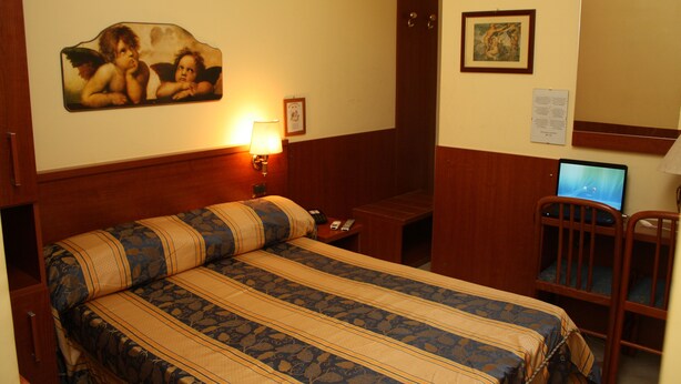 Quarto de hotel com uma cama, uma escrivaninha, uma cadeira, uma luminária e obras de arte nas paredes.