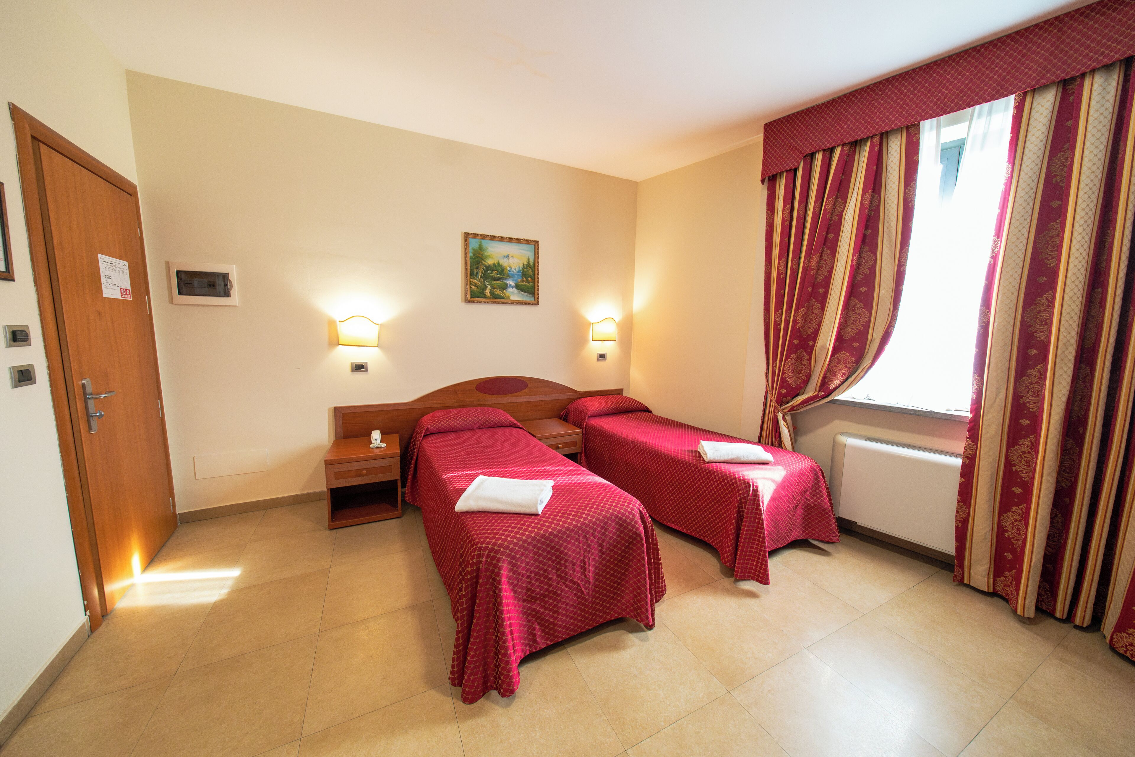 Quarto de hotel com duas camas de solteiro, cabeceira de madeira, um quadro na parede e uma janela com cortinas.