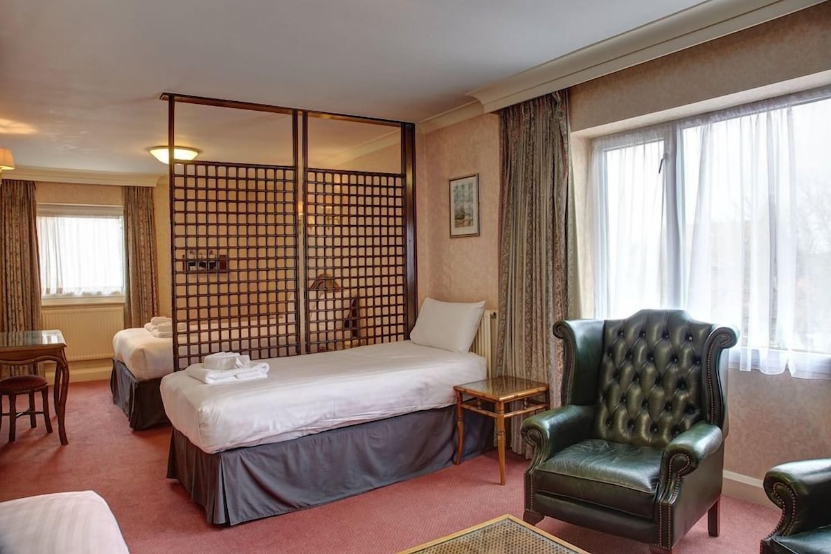 Quarto de hotel com cama, cadeira, mesinha, escrivaninha e janela com cortinas.