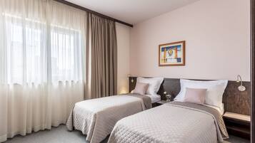 Quarto de hotel com duas camas, roupa de cama bege, um quadro na parede e uma janela com cortinas.
