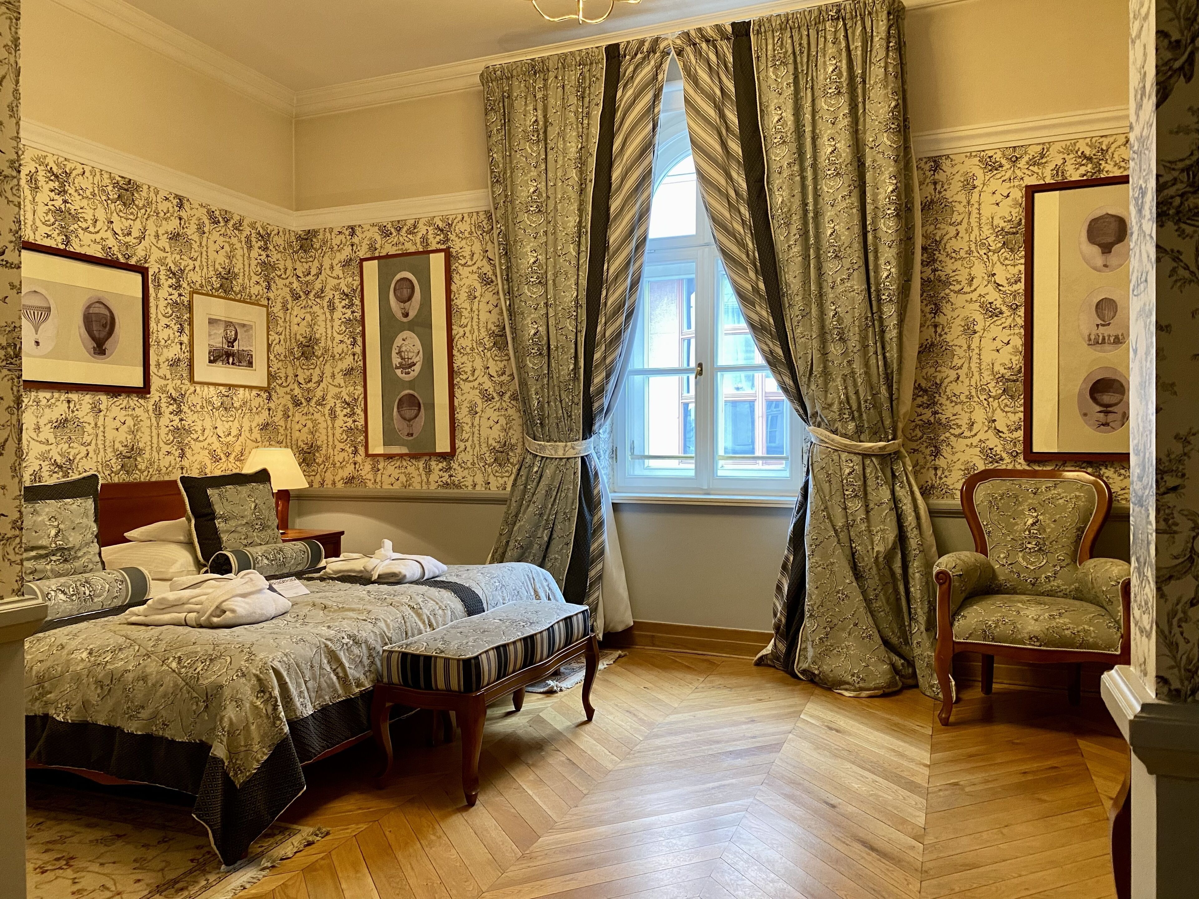 Quarto tradicional com piso de madeira, cama com colcha florida, uma cadeira e cortinas.