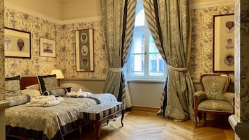 Quarto tradicional com piso de madeira, cama com colcha florida, uma cadeira e cortinas.