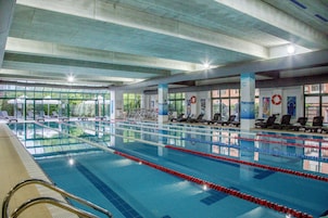 Piscina interior com várias pistas de natação, espreguiçadeiras e vista para o exterior através de grandes janelas.
