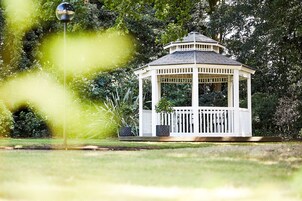 Um gazebo com exterior branco e teto escuro, rodeado por vegetação exuberante.