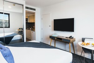 Quarto de hotel moderno com uma cama grande, uma mesa de escritório com televisão de tela plana, uma pequena mesa de jantar com doces e uma kitchenette ao fundo.
