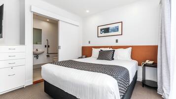 Quarto de hotel com uma cama grande, mesa de cabeceira com telefone, espelho, banheiro com pia e vaso sanitário, e um quadro na parede.