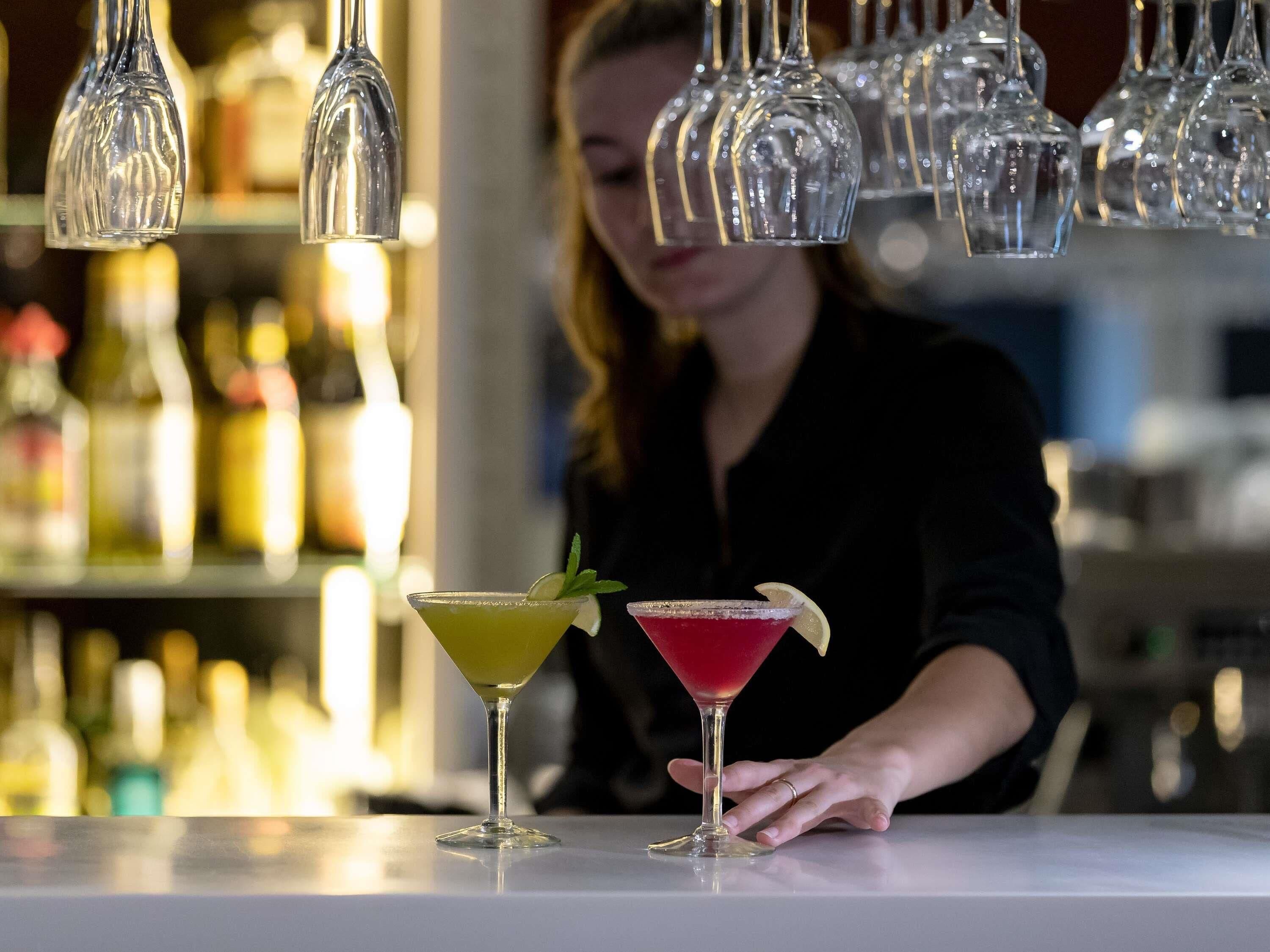 Um bar com dois drinks, um bartender e prateleiras com garrafas ao fundo.