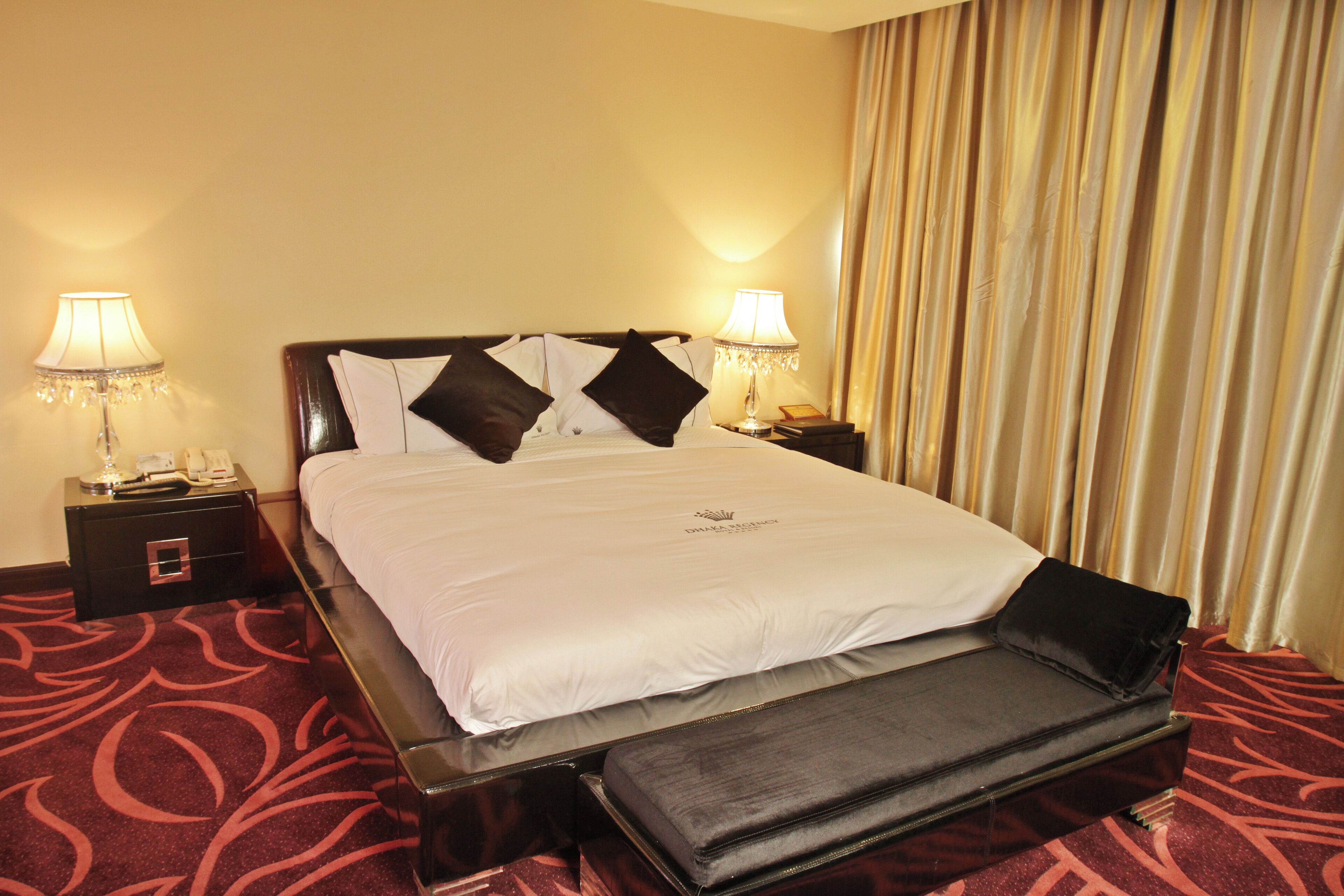 Quarto de hotel com uma cama grande, duas mesas de cabeceira com luminárias, um banco aos pés da cama e um tapete estampado em vermelho.