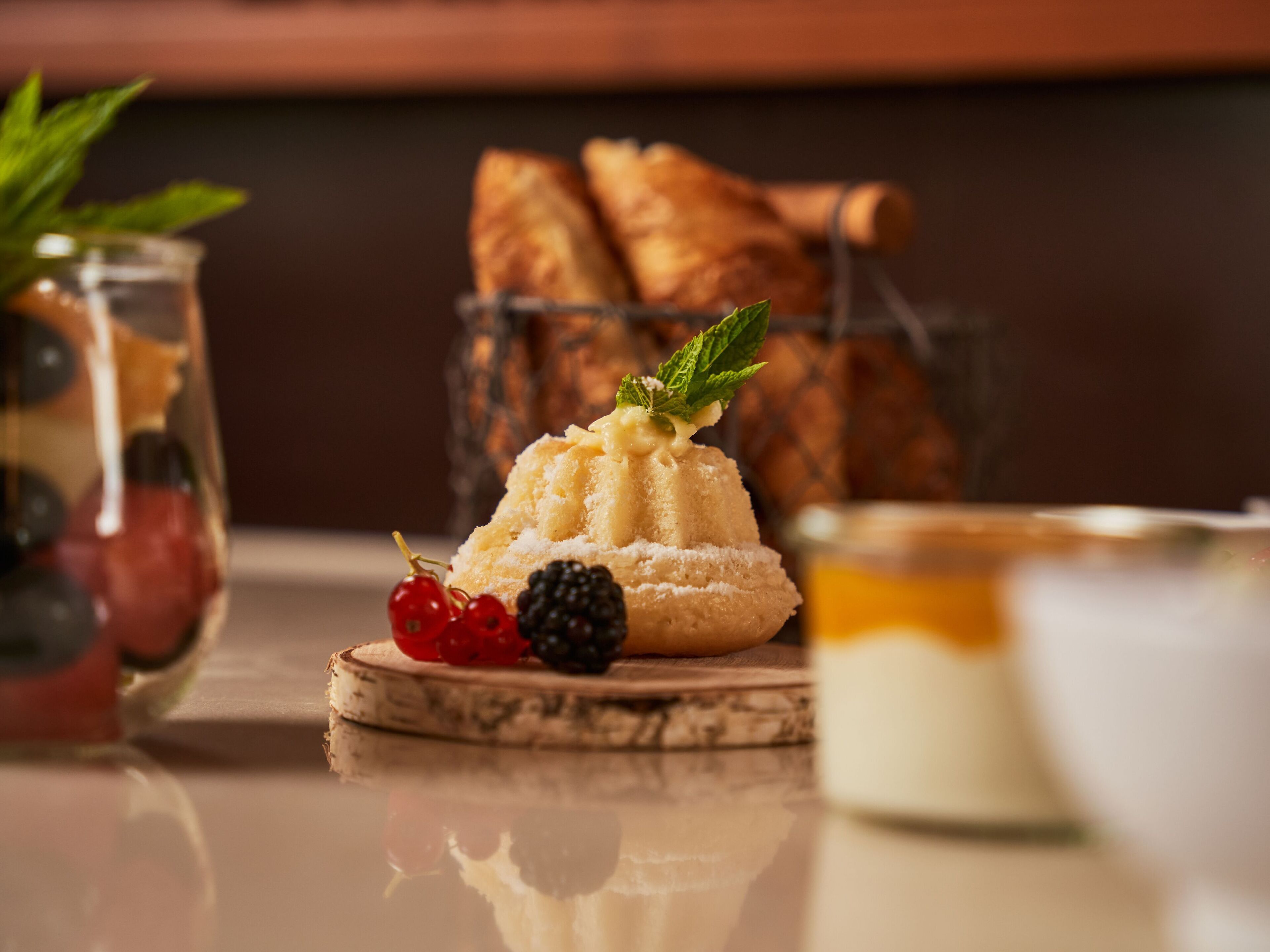 Sobremesa com frutos vermelhos e hortelã sobre uma tábua de madeira, com doces e um pote ao fundo.