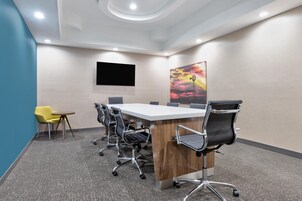 Uma sala de conferências com uma mesa comprida, seis cadeiras, uma televisão e um quadro colorido na parede.