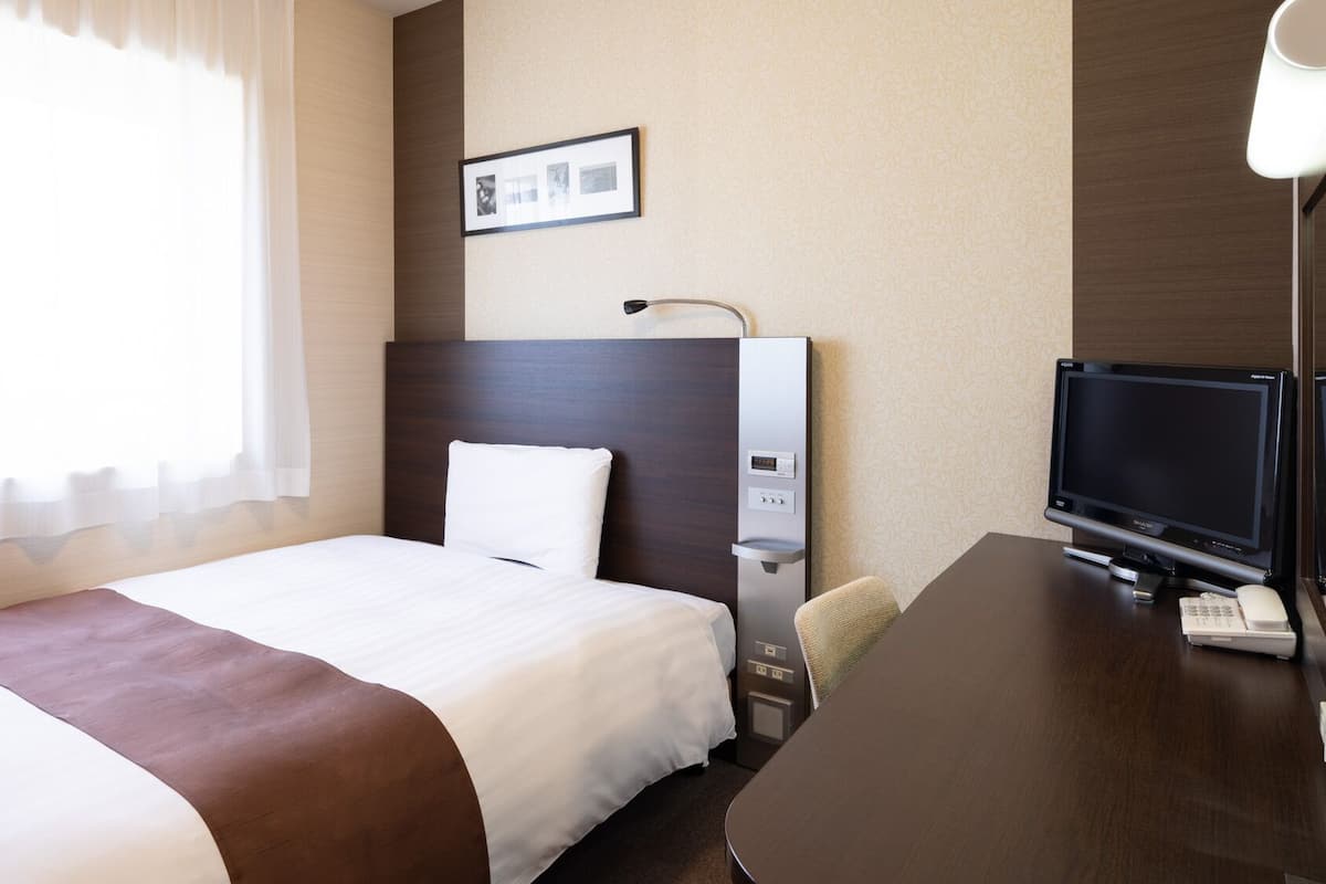 Quarto de hotel com cama, escrivaninha com televisão, luminária de mesa, quadro na parede e janela com cortinas.