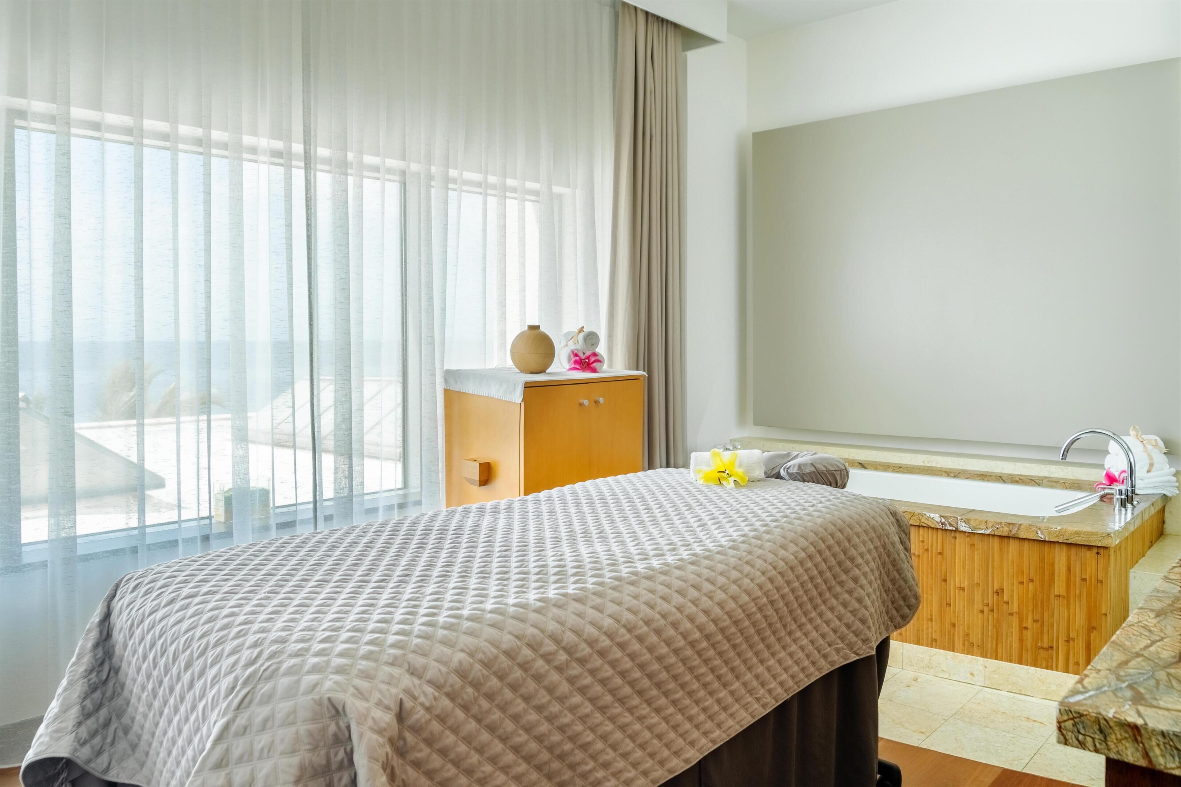 Quarto de hotel com cama, armário de madeira, banheiro com banheira e uma janela grande com cortinas.