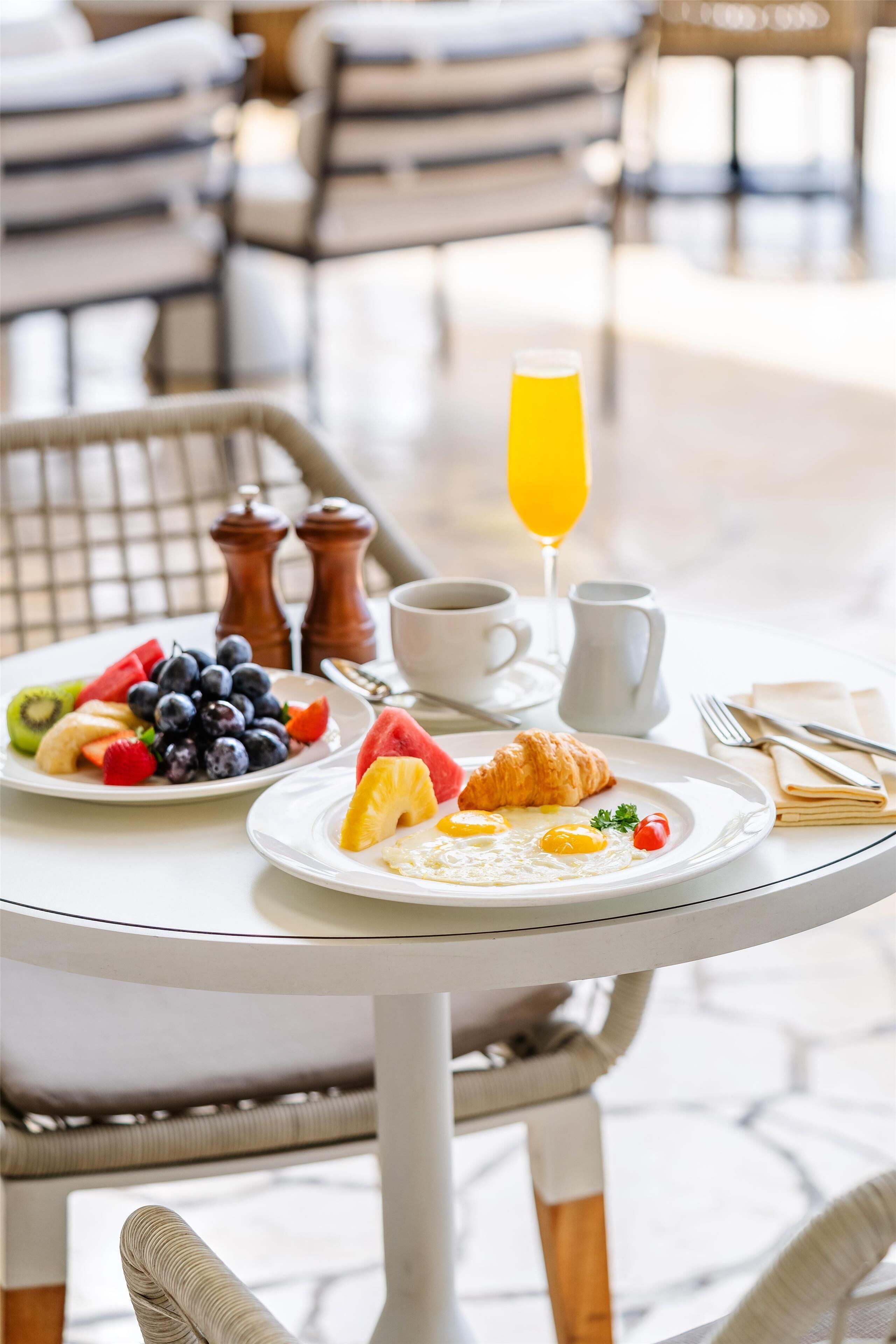 Mesa redonda com pratos de fruta, ovos e croissants, um copo de sumo de laranja e uma chávena de café.