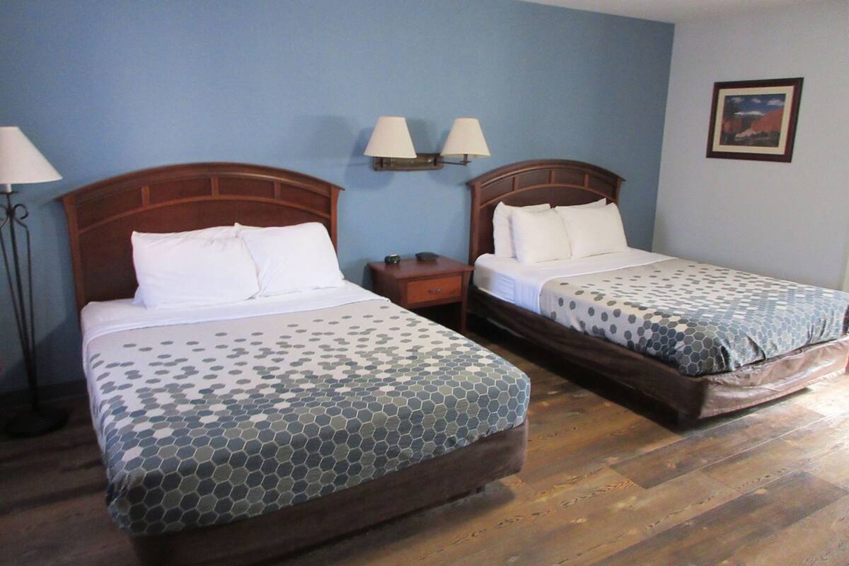 Duas camas de casal com cabeceiras de madeira, uma mesinha de cabeceira, duas luminárias fixadas na parede e um quadro emoldurado na parede.