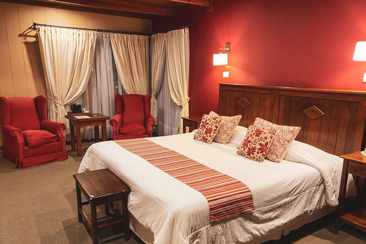 Quarto de hotel com uma cama grande, duas poltronas vermelhas, uma mesa de cabeceira de madeira e uma pequena mesa.