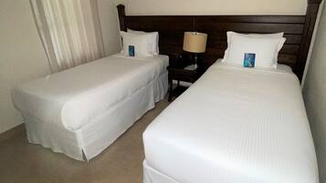 Quarto de hotel com duas camas de solteiro, uma mesa de cabeceira com abajur, um relógio de parede e uma obra de arte emoldurada.