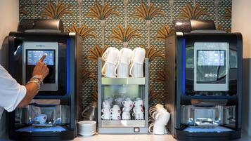 Uma estação de café com duas máquinas de café KREA, uma seleção de chávenas brancas e uma pilha de pratos.