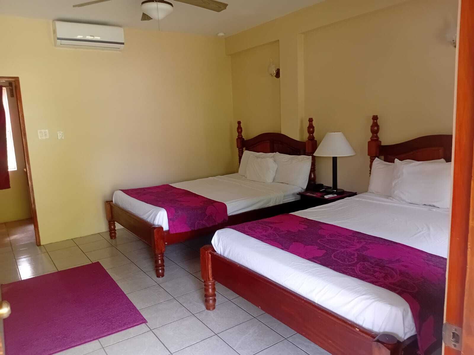 Quarto de hotel com duas camas de madeira, cada uma com colchão branco e cobre roxo. Há um ventilador de teto, uma mesa de cabeceira com abajur e uma janela com cortinas.