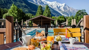 Mesa de café da manhã com croissants, frutas, suco e um jornal, com vista para uma piscina e montanhas.