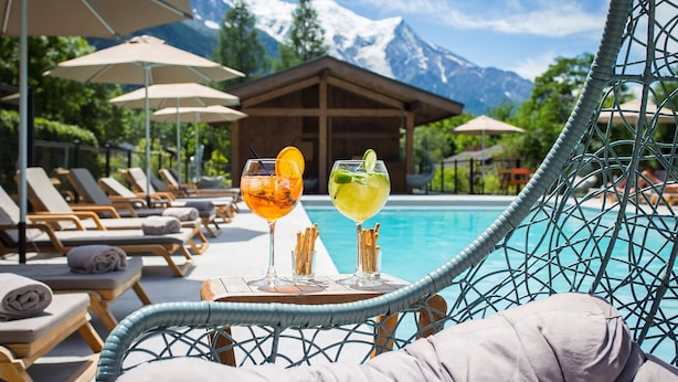 Uma área de descanso à beira da piscina, com duas bebidas sobre uma mesa, cadeiras de descanso e vista para as montanhas.
