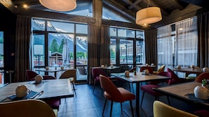 Restaurante com grandes janelas oferecendo vista para as montanhas, mesas de madeira e cadeiras vermelhas.