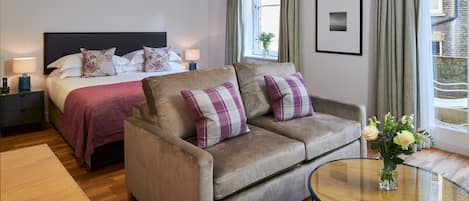 Quarto de hotel com cama, sofá, mesa de vidro e janela com cortinas.