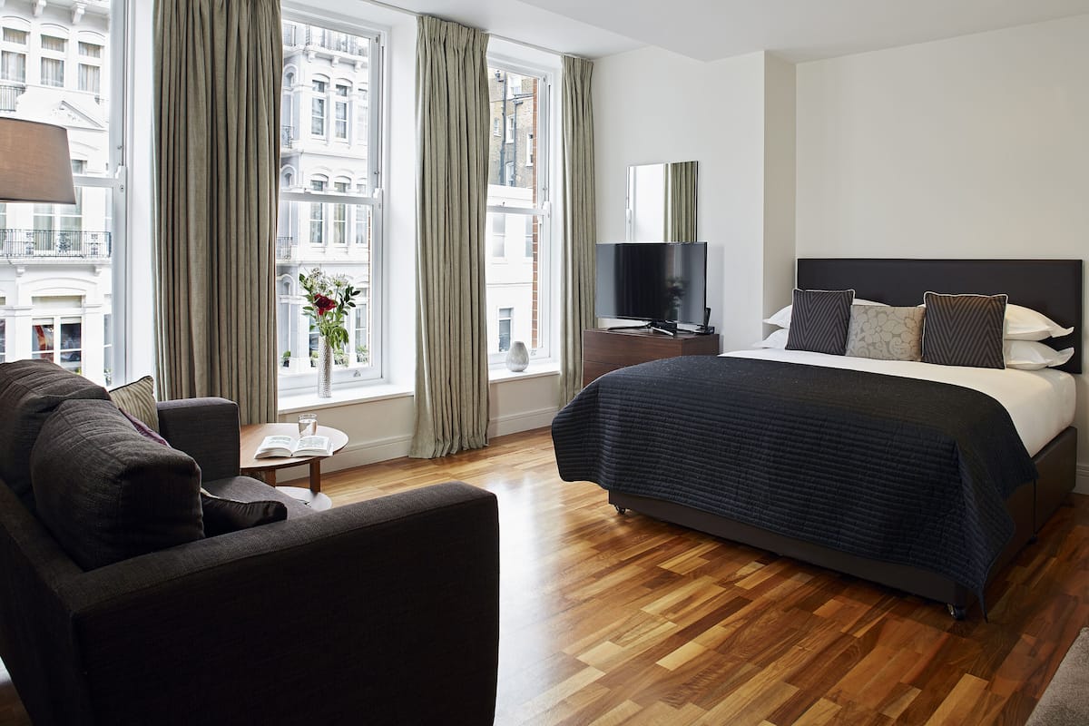Quarto de hotel com cama, televisão, sofá, mesa pequena e janela com cortinas.