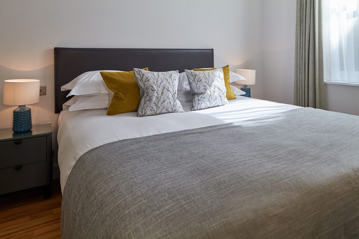 Cama bem arrumada com manta cinza, almofadas brancas e duas almofadas amarelo-mostarda. Há duas mesas de cabeceira com luminárias e uma janela com cortinas.
