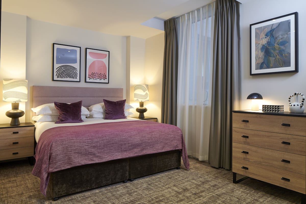 Quarto de hotel com cama, duas mesas de cabeceira com abajures, cômoda e obras de arte nas paredes.
