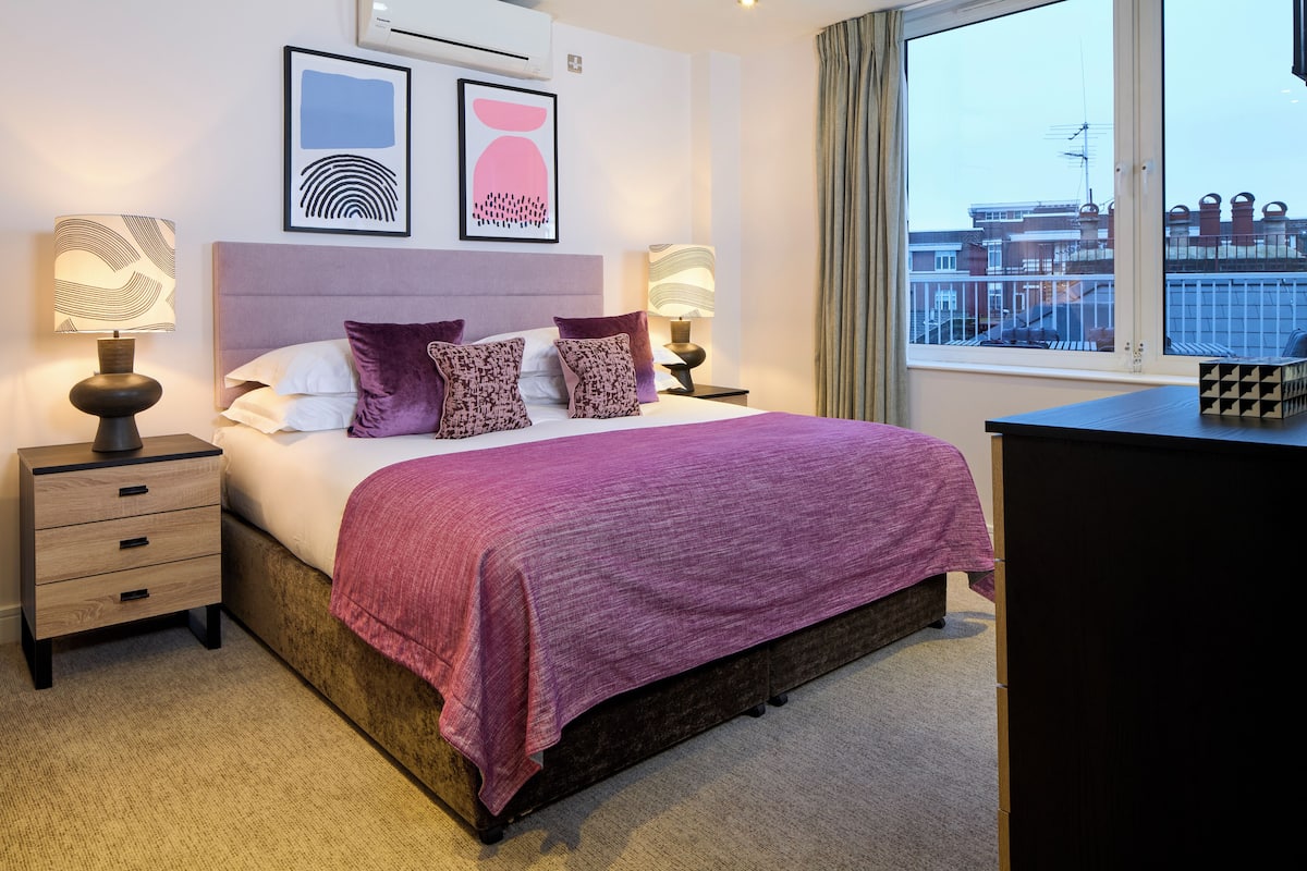 Quarto de hotel moderno com uma cama grande, duas mesas de cabeceira com luminárias, uma cômoda e vista para a cidade através de uma janela ampla.