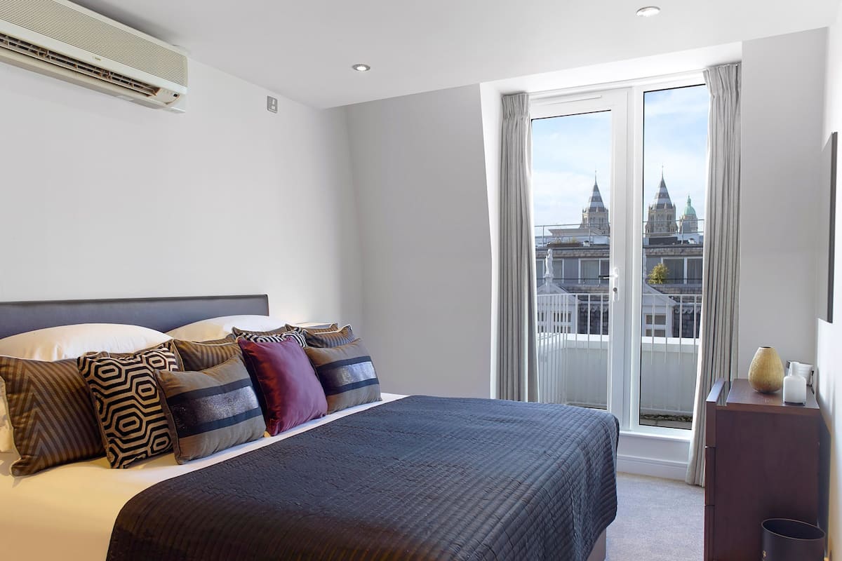 Quarto de hotel com uma cama grande, vista para a paisagem urbana através de uma porta deslizante, uma mesa pequena com um vaso e uma lixeira.