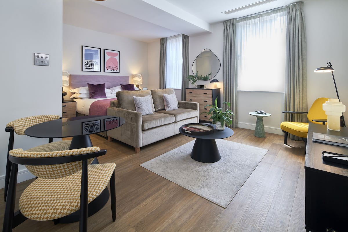 Quarto de hotel moderno com cama, sofá, mesa de jantar, cadeiras e uma pequena mesa lateral com luminária.