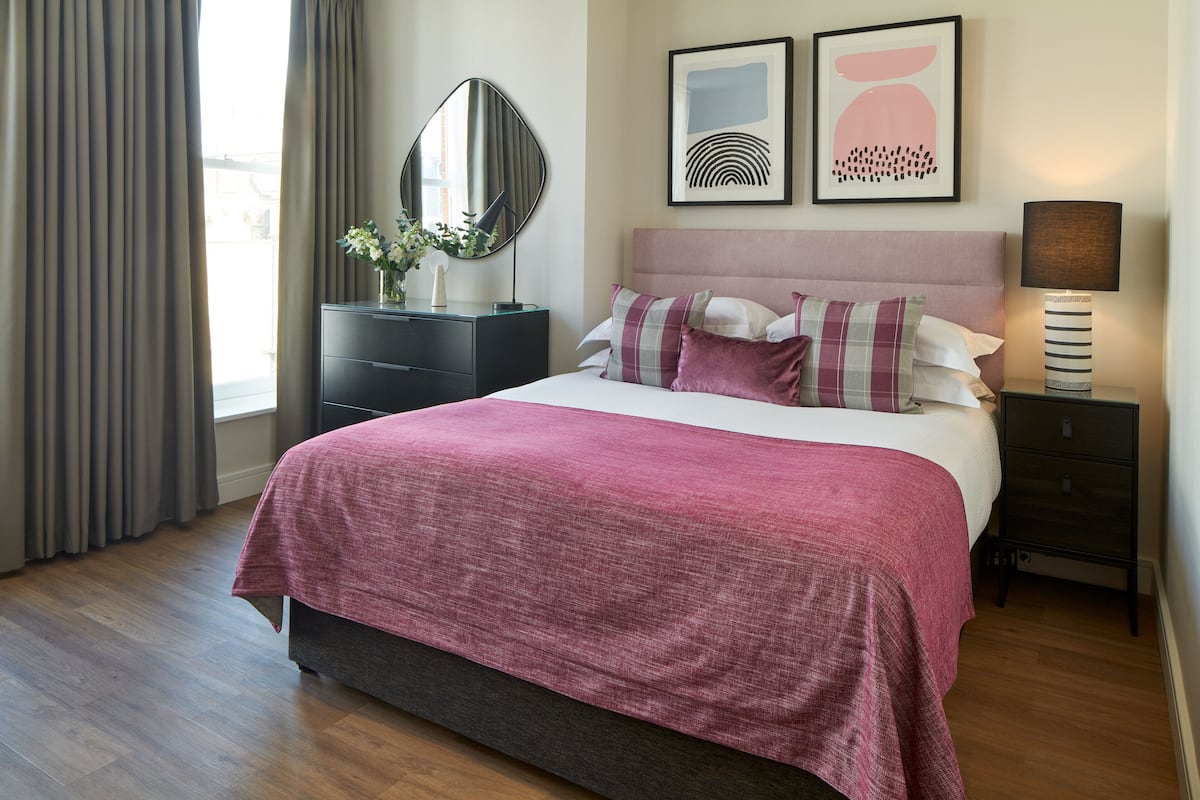 Quarto com cama, penteadeira, espelho, mesa de cabeceira com abajur e duas obras de arte emolduradas na parede.