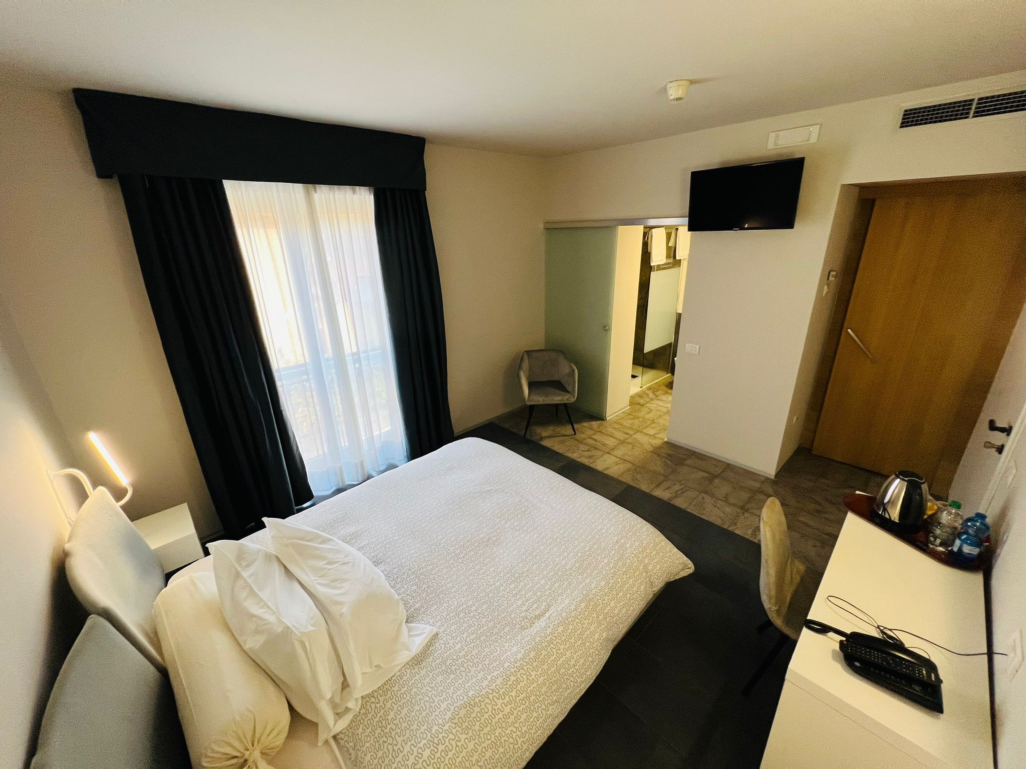 Quarto de hotel com cama, escrivaninha, cadeira, televisão e banheiro visível através de uma porta aberta.