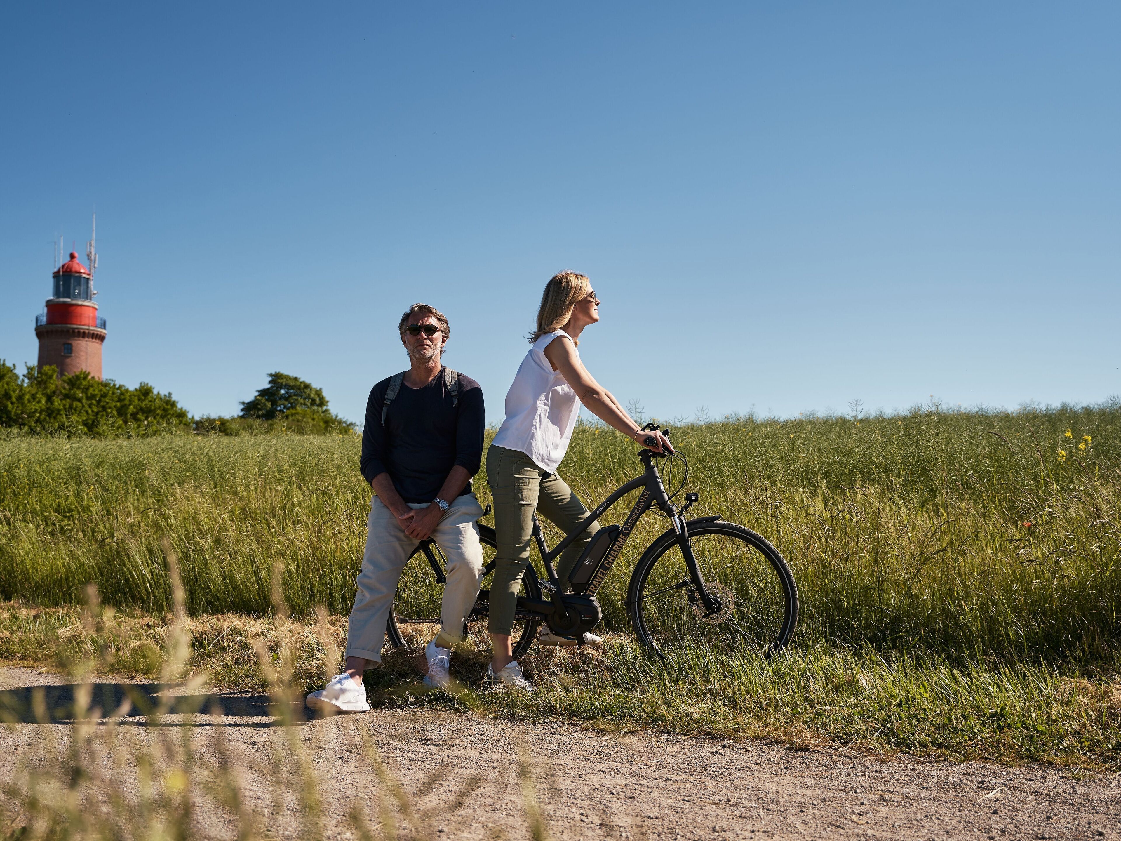 Duas pessoas em um caminho de terra, com uma bicicleta, e um farol ao fundo, além de um campo gramado.