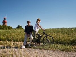 Duas pessoas em um caminho de terra, com uma bicicleta, e um farol ao fundo, além de um campo gramado.