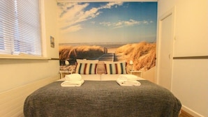 Quarto com mural de tema praiano, cama com duas almofadas e janela com persianas.