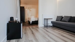 Quarto de hotel moderno com sofá cinzento, uma mesa pequena e um aquecedor compacto preto.