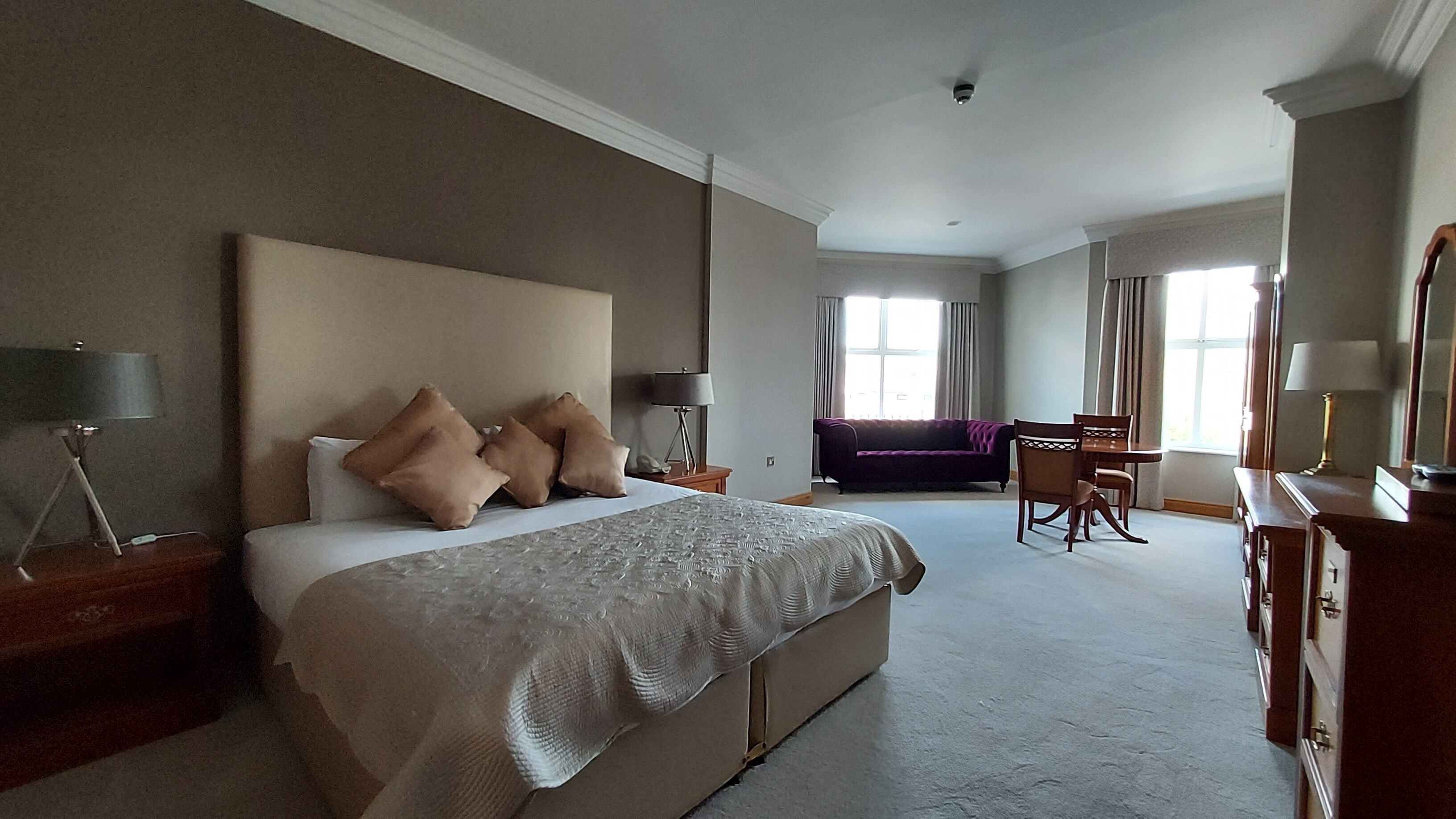 Quarto de hotel com uma cama grande, mesas de cabeceira com luminárias, uma escrivaninha com cadeira e um sofá roxo.