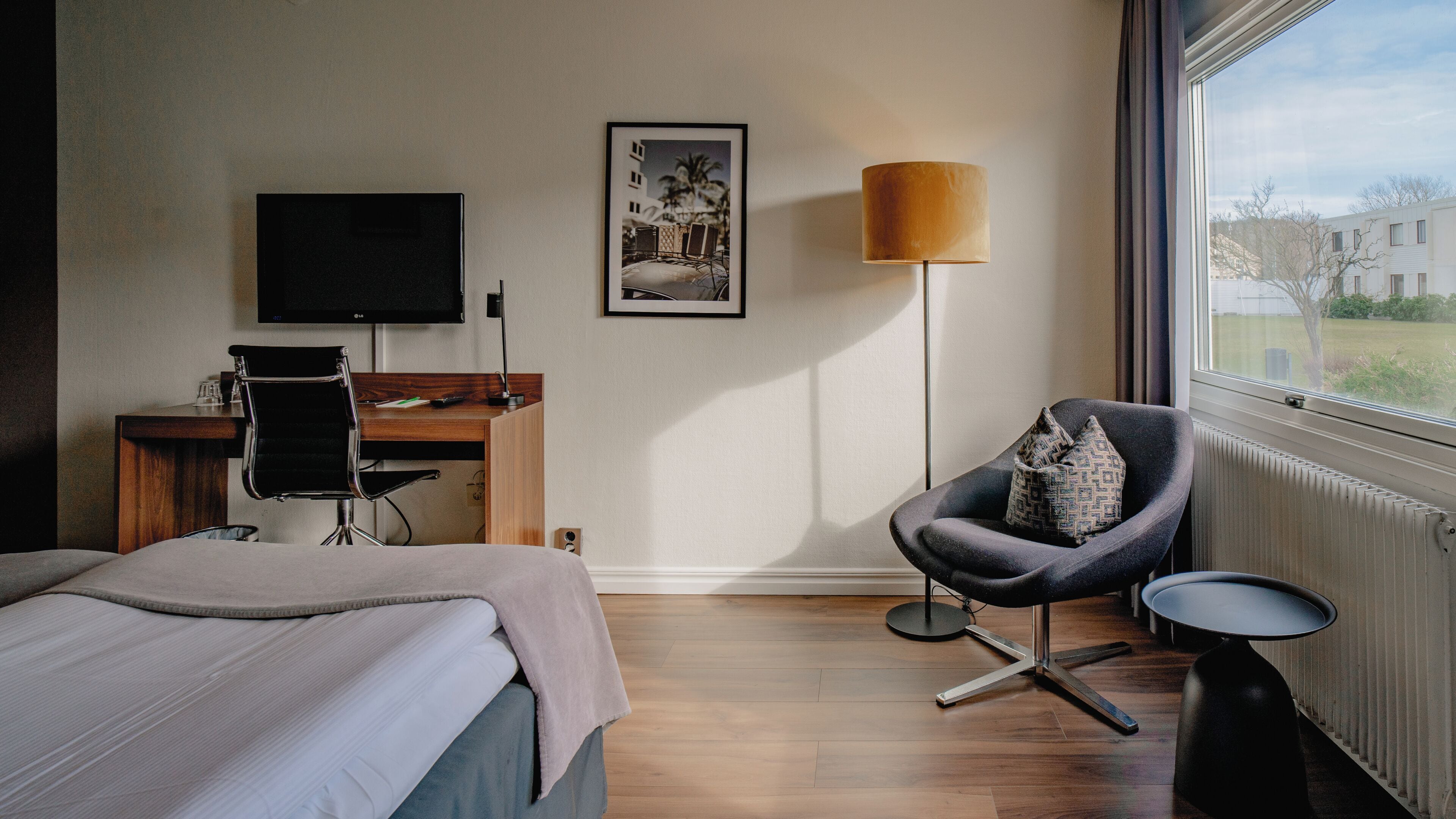 Quarto de hotel com cama, escrivaninha, cadeira, televisão, luminária e janela com vista.