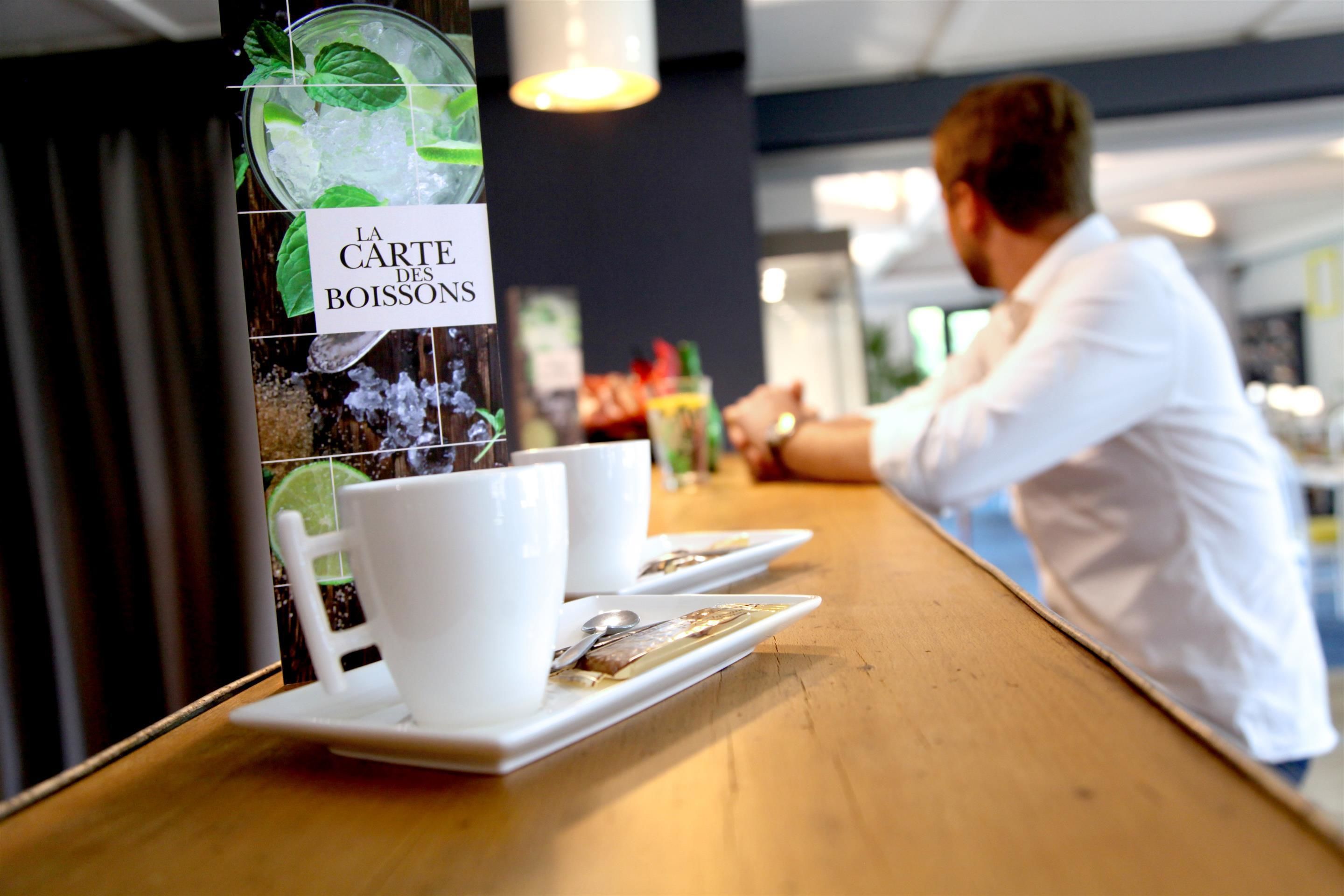 Um balcão de café com um quadro de menu exibindo 'LA CARTE DES BOISSONS' e duas chávenas brancas numa bandeja.