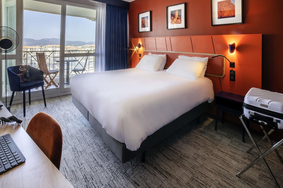 Quarto de hotel com uma cama grande, uma escrivaninha com teclado, uma cadeira, uma mala e uma varanda com vista para a cidade.