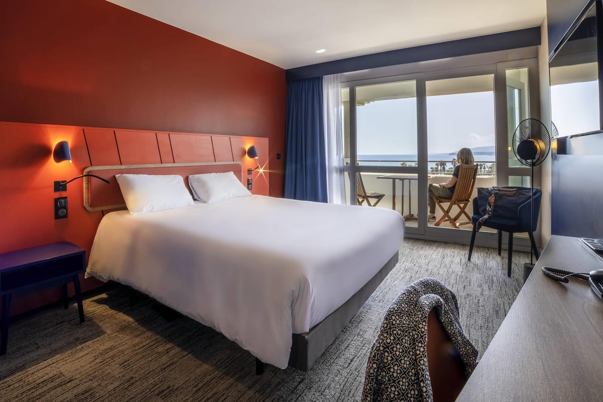 Quarto de hotel com cama, mesas de cabeceira, escrivaninha, cadeira e vista para o mar.