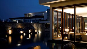 Um edifício moderno com fachada de vidro, piscina com pedras e interior bem iluminado com mobiliário.