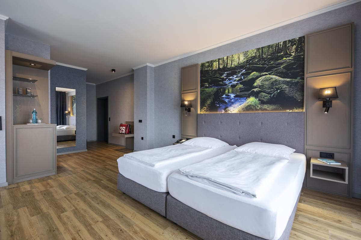 Quarto de hotel moderno com uma cama grande, banheiro com espelho e televisão na parede exibindo uma paisagem natural.