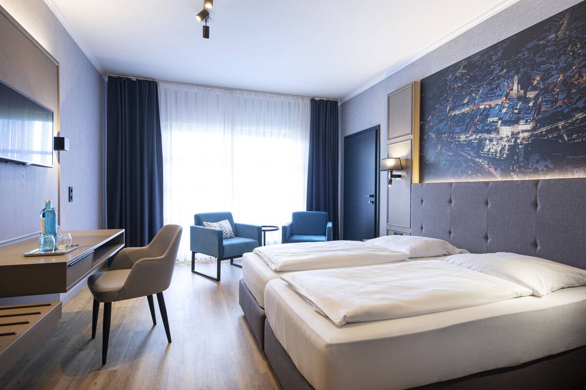 Quarto de hotel com uma cama, duas cadeiras, uma escrivaninha e uma janela grande com cortinas.